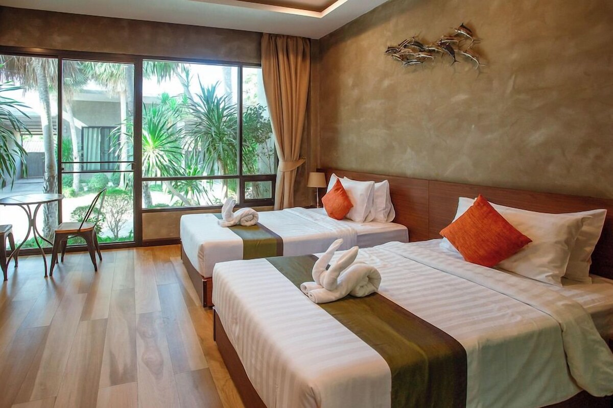 Quarto de hotel com duas camas, uma mesinha e vista para palmeiras através de grandes janelas.