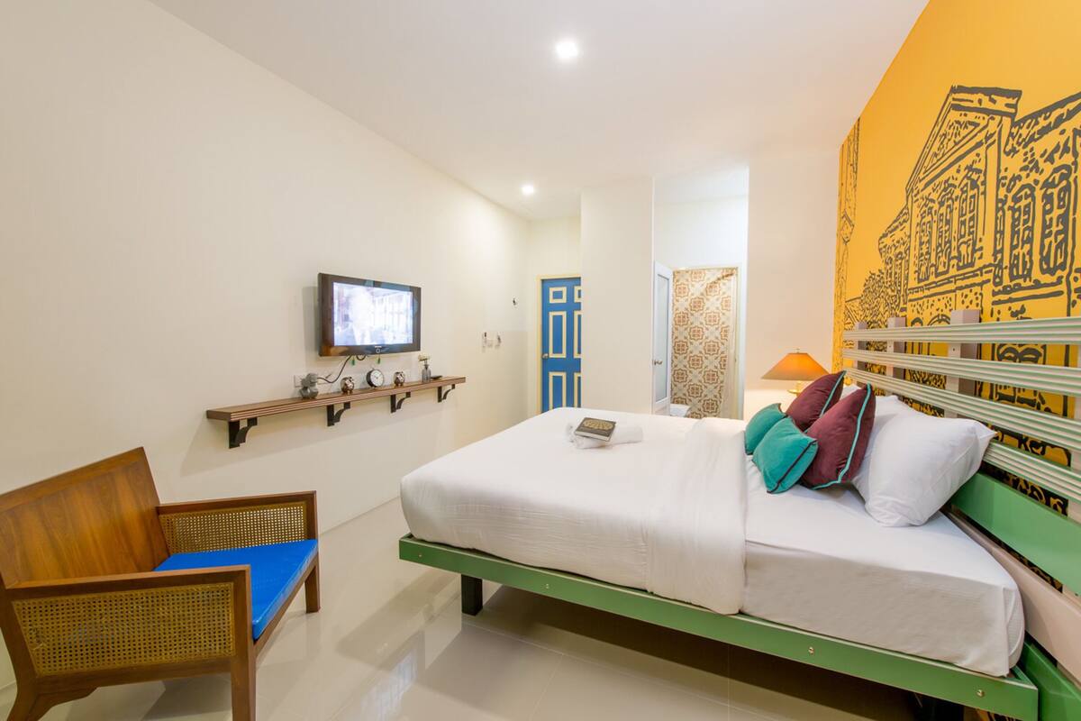 Quarto de hotel com cama, televisão, cadeira e parede com um mural.