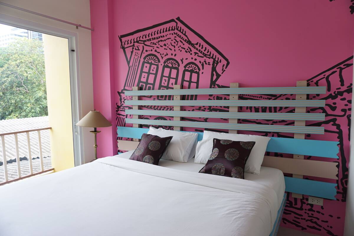 Uma cama com roupa de cama branca, uma parede rosa com um mural de um edifício preto e uma janela com vista para árvores.