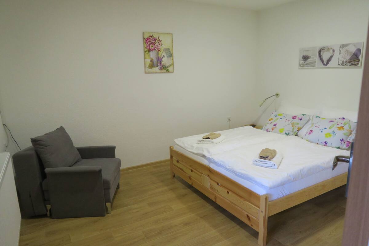 Quarto com cama de madeira, sofá cinzento e uma pequena mesa.