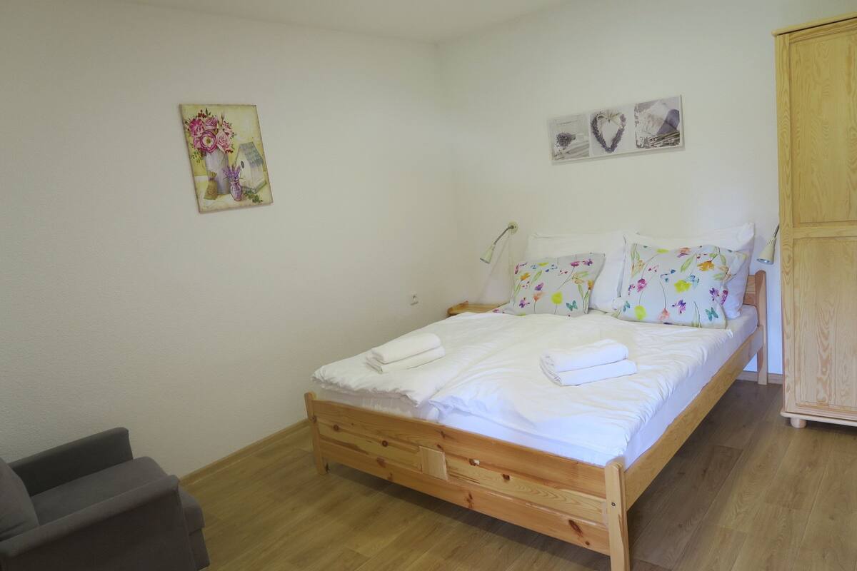 Uma cama bem arrumada com lençóis brancos e cabeceira de madeira, uma pequena mesa de cabeceira com abajur e um guarda-roupa de madeira.