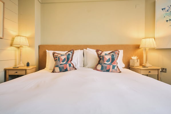 Uma cama bem arrumada com almofadas estampadas, duas luminárias de cabeceira, um criado-mudo com uma lâmpada e uma obra de arte pendurada na parede.