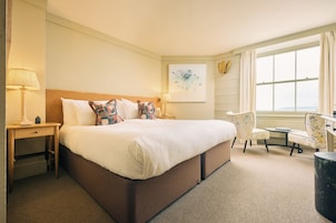 Quarto de hotel com uma cama grande, mesas de cabeceira, uma cadeira, uma pequena mesa e uma janela com cortinas.