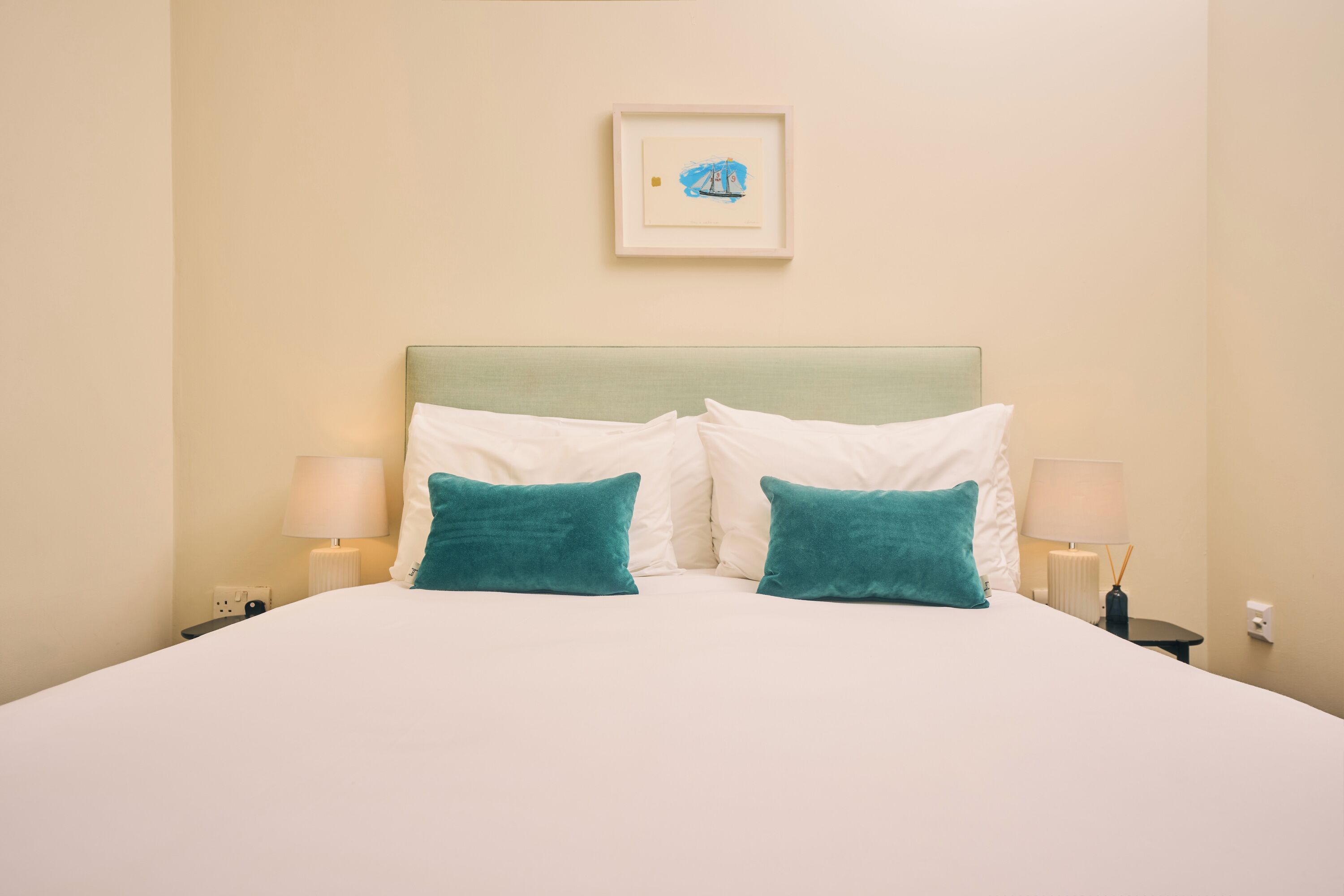 Cama bem arrumada com almofadas brancas e azul-turquesa, um quadro na parede e duas luminárias de cabeceira.