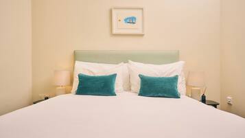 Cama bem arrumada com almofadas brancas e azul-turquesa, um quadro na parede e duas luminárias de cabeceira.