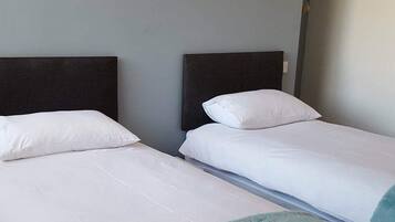 Quarto de hotel com duas camas, cada uma com roupa de cama branca e manta azul-esverdeada. Existem luminárias fixadas na parede, de cada lado das camas.