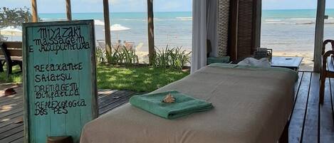 Um deck de madeira com uma mesa de massagem, uma placa listando os serviços e vista para a praia.