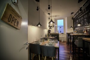Uma área de refeições com mesa de madeira posta para quatro, cadeiras, luminárias pendentes e um letreiro na parede com a inscrição 'WELCOME OPEN'.