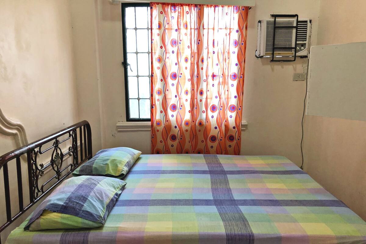 Uma cama de solteiro com um edredom colorido xadrez e duas almofadas, uma janela com padrão quadriculado e um ar condicionado na parede.