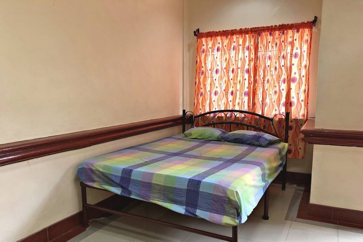 Quarto pequeno com uma cama de solteiro, estrutura de metal, colcha xadrez colorida e cortina laranja estampada.
