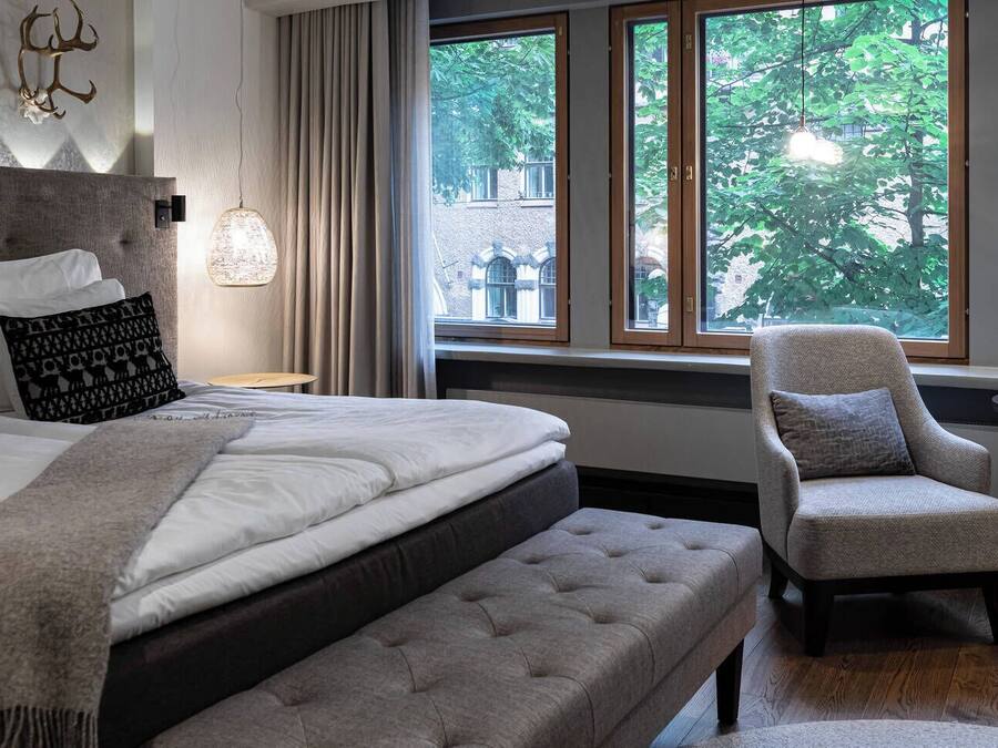 Quarto de hotel moderno com uma cama grande, uma poltrona cinza, uma pequena mesa e vista para o verde lá fora.
