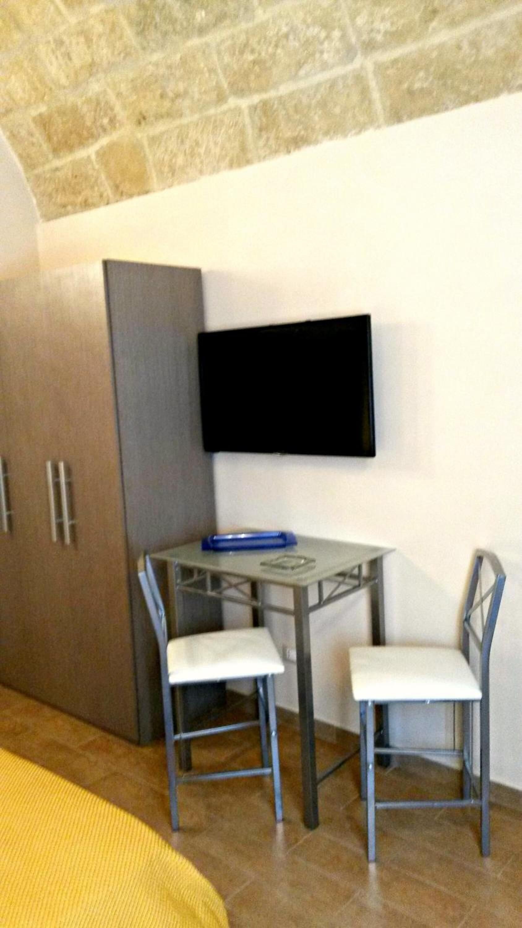 Quarto pequeno com cama, escrivaninha com duas cadeiras, televisão de tela plana na parede e guarda-roupa.