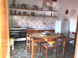 Cozinha com fogão, forno, geladeira e mesa de jantar com cadeiras. Há prateleiras acima do balcão com diversos utensílios de cozinha e um relógio na parede.