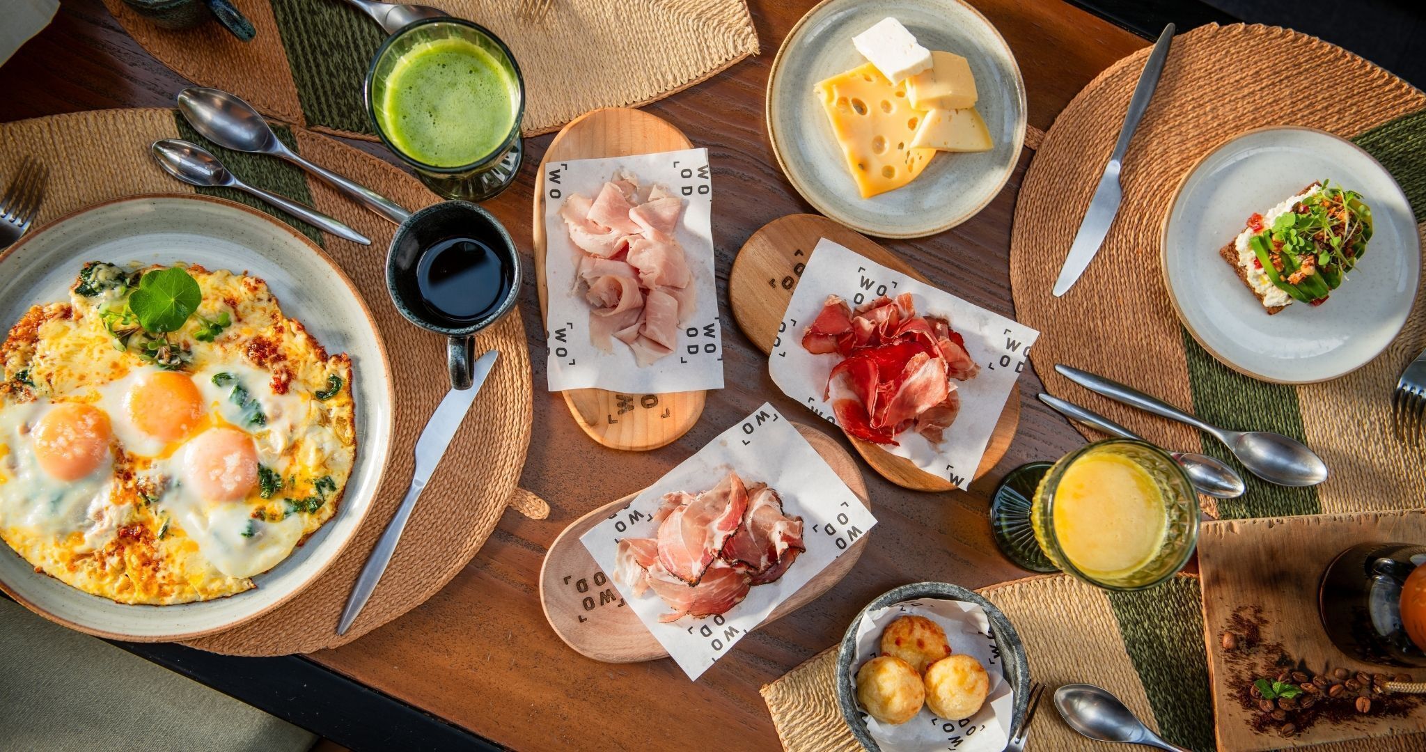 Disposição de café da manhã com diversos pratos, incluindo ovos, queijo e carnes, sobre uma mesa de madeira com jogos americanos trançados.