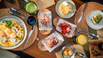 Disposição de café da manhã com diversos pratos, incluindo ovos, queijo e carnes, sobre uma mesa de madeira com jogos americanos trançados.
