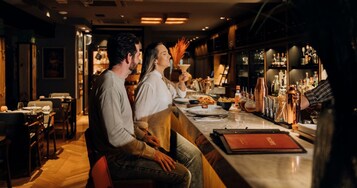 Uma área de bar com balcão, banquetas e diversos tipos de garrafas e copos.