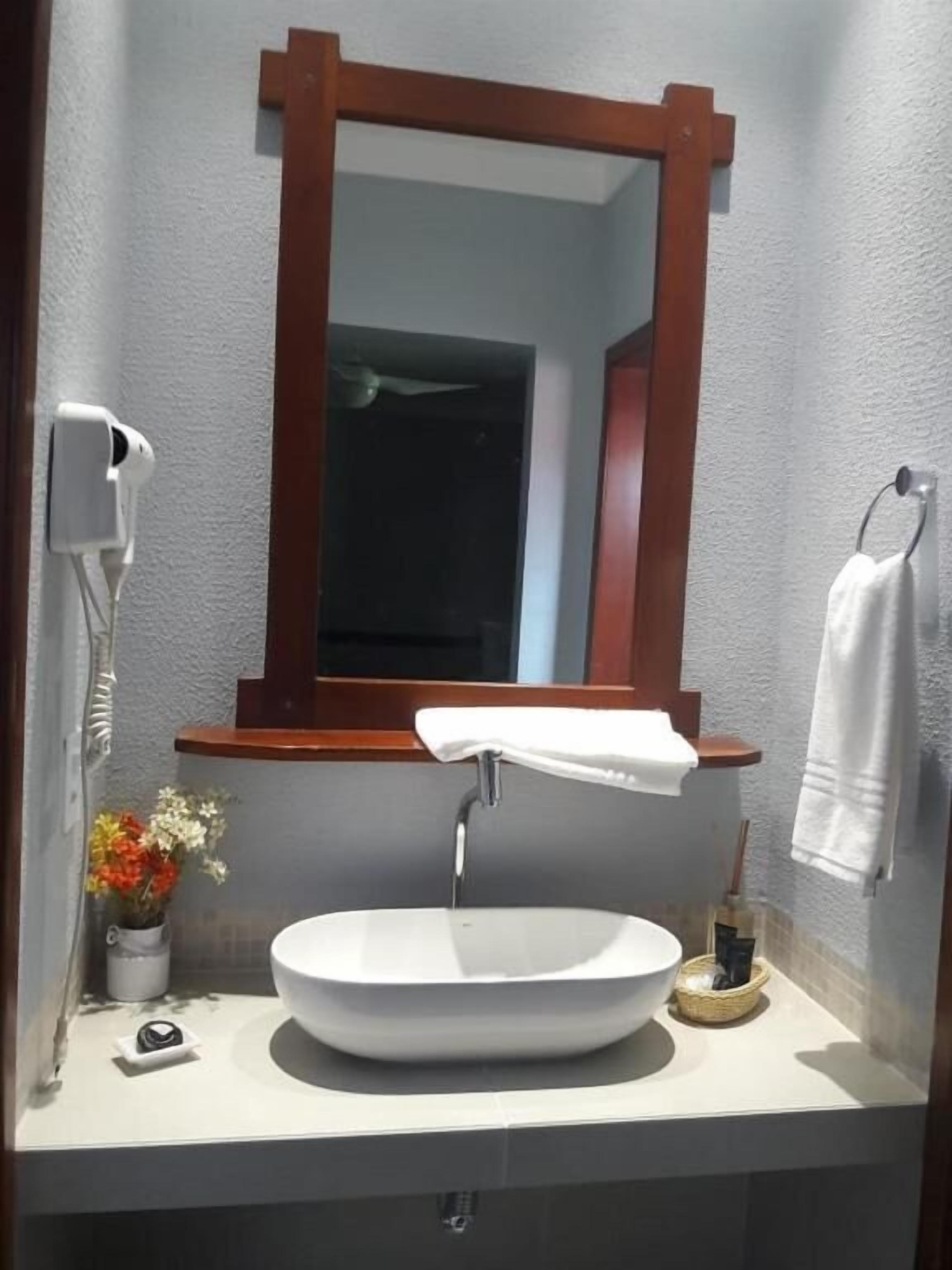 Casa de banho com lavatório branco, espelho com moldura de madeira, secador de cabelo e toalheiro.