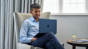 Uma pessoa sentada em uma cadeira com um laptop, um copo de água e uma revista sobre uma mesinha.