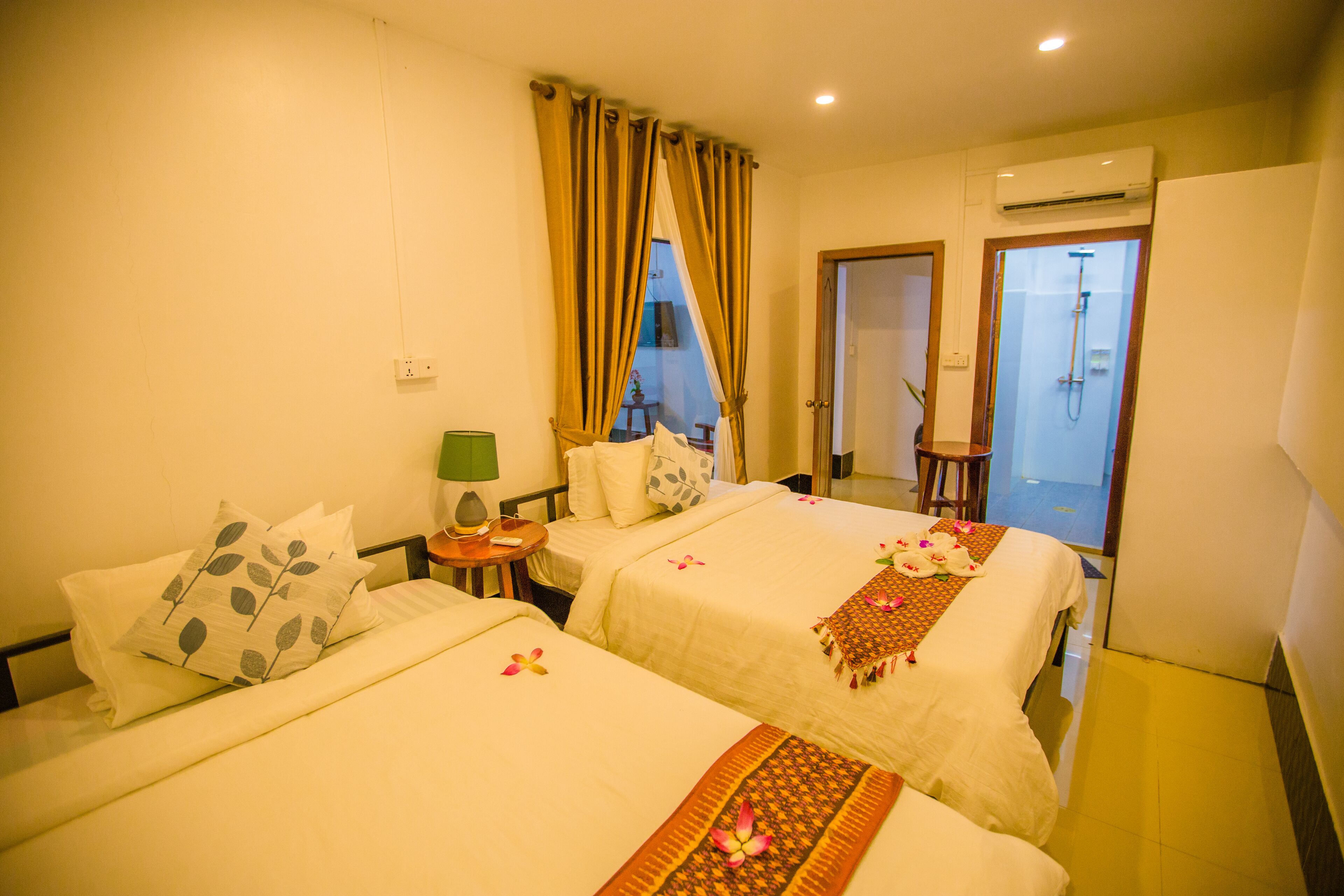 Quarto de hotel com duas camas, uma mesinha com luminária, janela com cortinas e aparelho de ar condicionado.