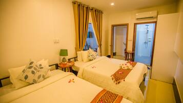 Quarto de hotel com duas camas, uma mesinha com luminária, janela com cortinas e aparelho de ar condicionado.