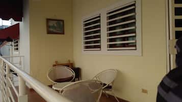 Uma varanda com grades de metal branco, duas cadeiras de vime, uma mesinha e uma janela com venezianas horizontais.