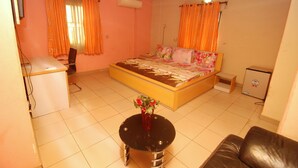 Um quarto com cama, escrivaninha, cadeira, mesinha de centro com um vaso, e um refrigerador.