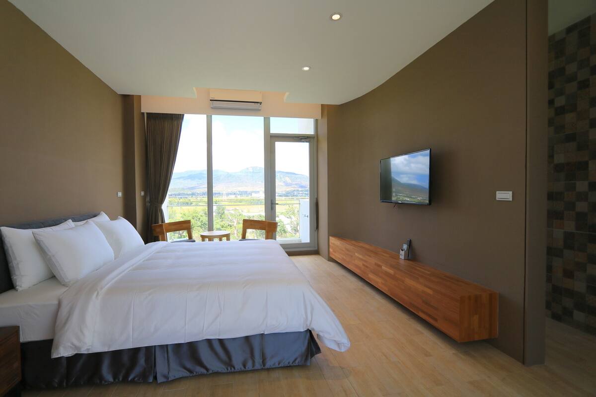 Quarto de hotel moderno com uma cama grande, um banco de madeira, uma TV e vista para as montanhas.