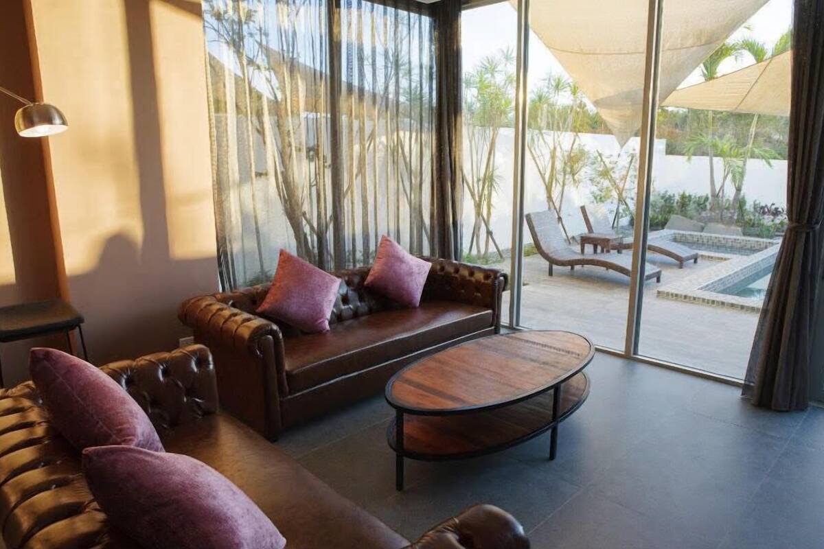 Uma sala de estar moderna com sofá de couro, mesa de centro de madeira e uma grande porta de vidro que dá para área externa com piscina e guarda-sóis.