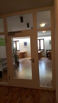 Um quarto com piso de madeira, porta com painel de vidro e vista para uma área de refeições com mesas e cadeiras.