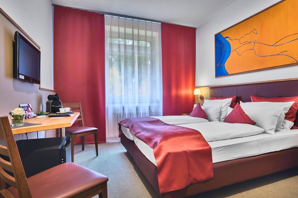 Quarto de hotel com cama, escrivaninha, cadeira, televisão e janela com cortinas.