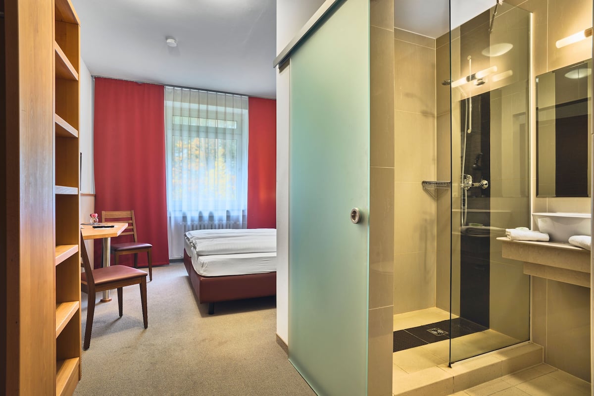 Quarto de hotel com cama, escrivaninha, cadeira, chuveiro e banheiro com paredes de vidro.