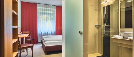 Quarto de hotel com cama, escrivaninha, cadeira, chuveiro e banheiro com paredes de vidro.