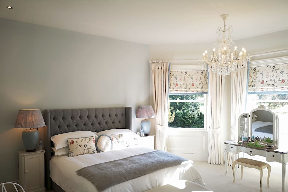Quarto com cabeceira estofada, cama grande com roupa de cama branca, um banquinho, mesa de cabeceira com abajur, cômoda e espelho.