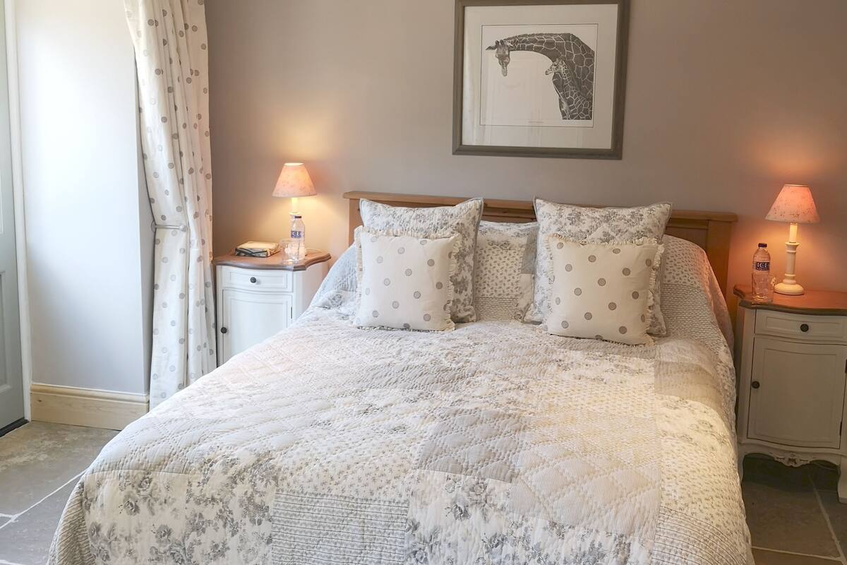 Cama bem arrumada com cobertura e almofadas estampadas em flor, flanqueada por duas mesas de cabeceira com abajures, uma ilustração emoldurada de uma girafa na parede e uma janela grande com cortinas.