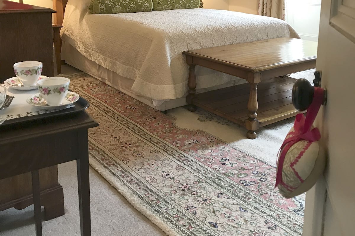 Quarto com cama, duas luminárias, mesa de cabeceira com xícaras, tapete e porta com sino.
