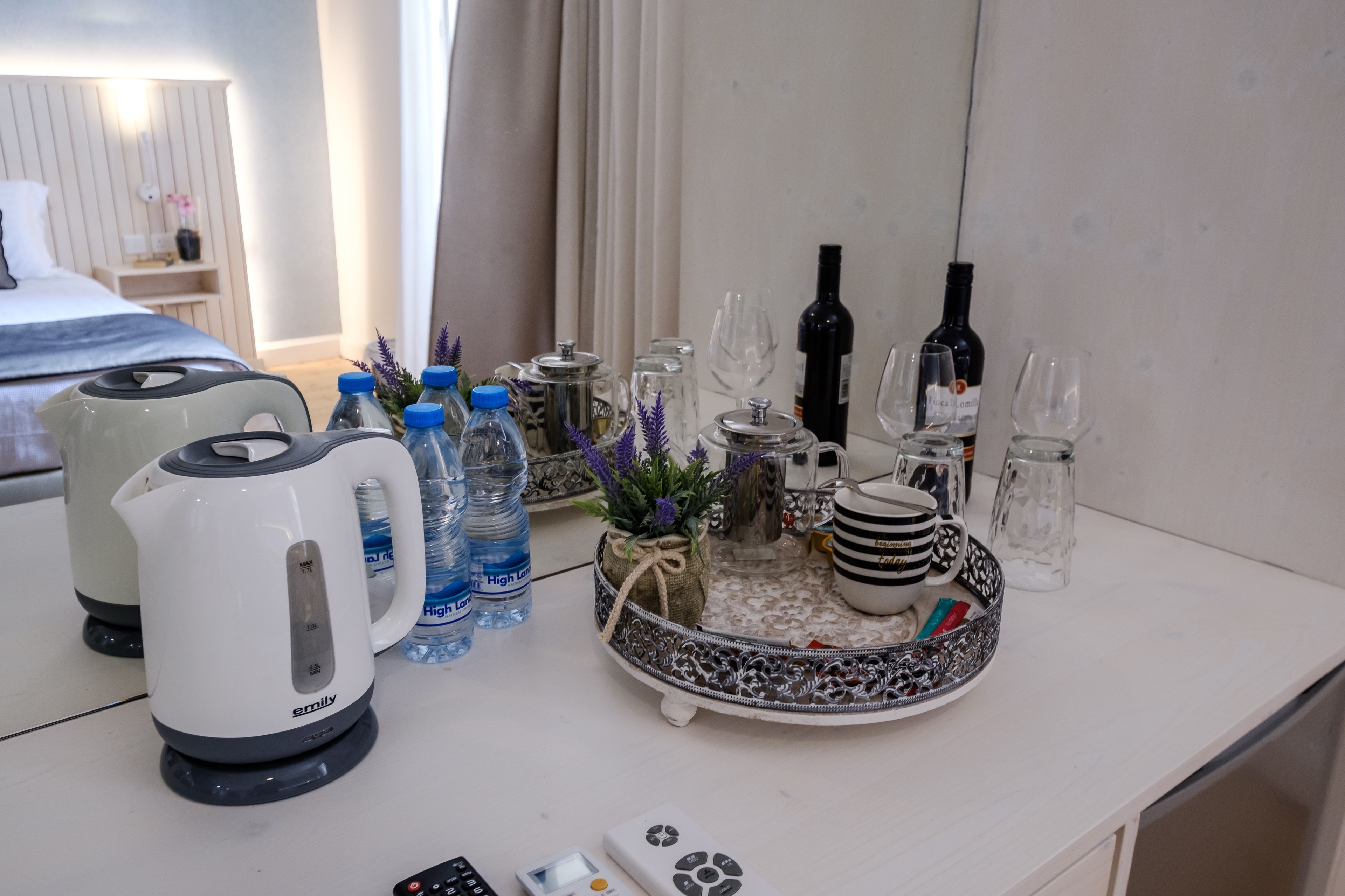 Quarto de hotel com bancada de cozinha, chaleira, garrafas de água, garrafas de vinho e bandeja com copos e uma pequena planta.