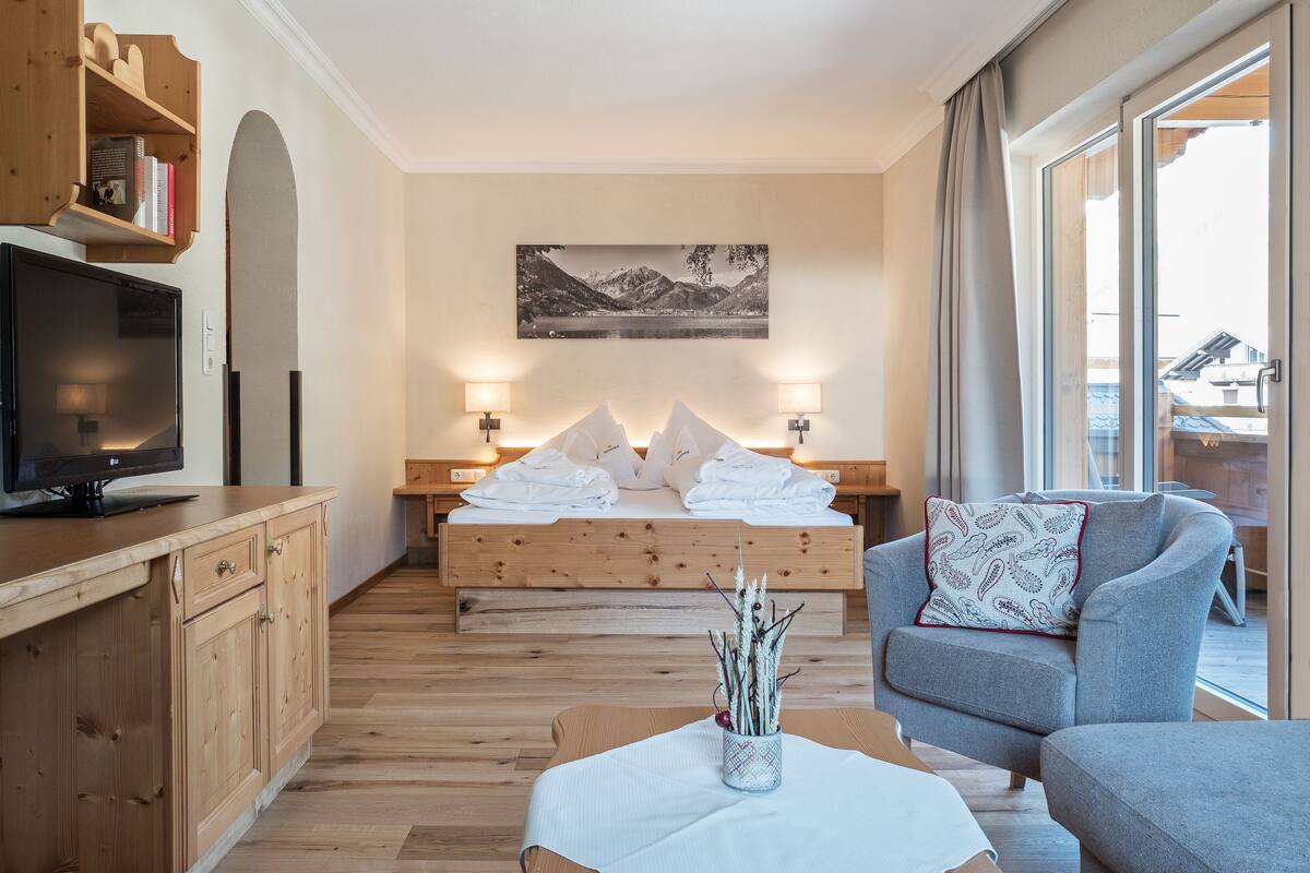 Quarto com cama, luminárias de cabeceira, televisão, sofá e janela com vista.