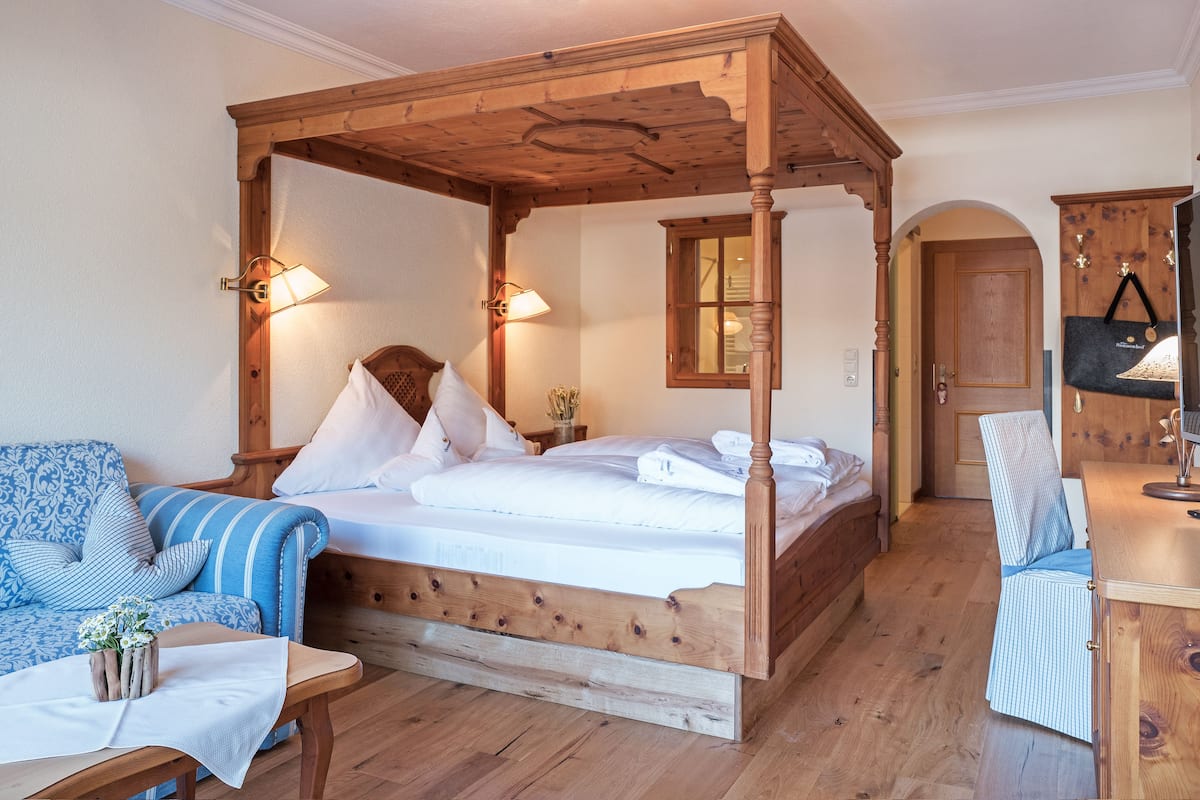 Uma cama de madeira com dossel, roupa de cama branca, um sofá listrado em azul e uma pequena mesa com um vaso de flores.