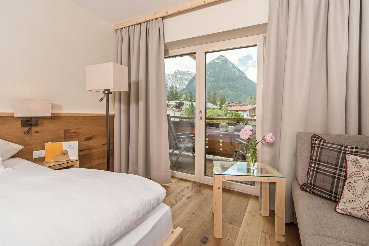 Quarto de hotel com cama, mesa de cabeceira com abajur, pequena mesa com um vaso de flores, sofá com almofada xadrez e porta de vidro deslizante que dá para uma varanda com vista para as montanhas.
