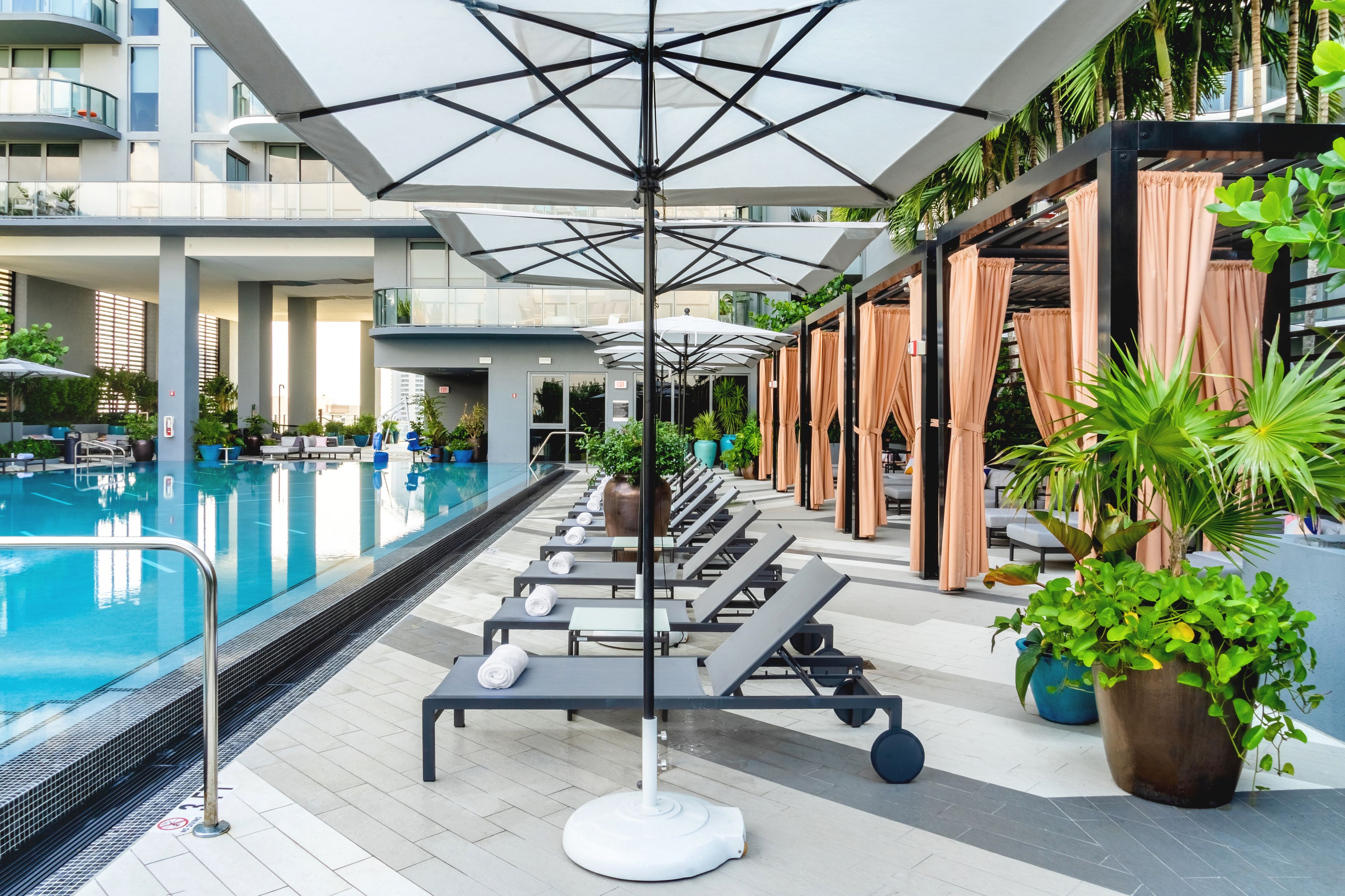 Uma área de piscina com espreguiçadeiras e guarda-sóis, rodeada por plantas em vasos e fachada de um edifício moderno.