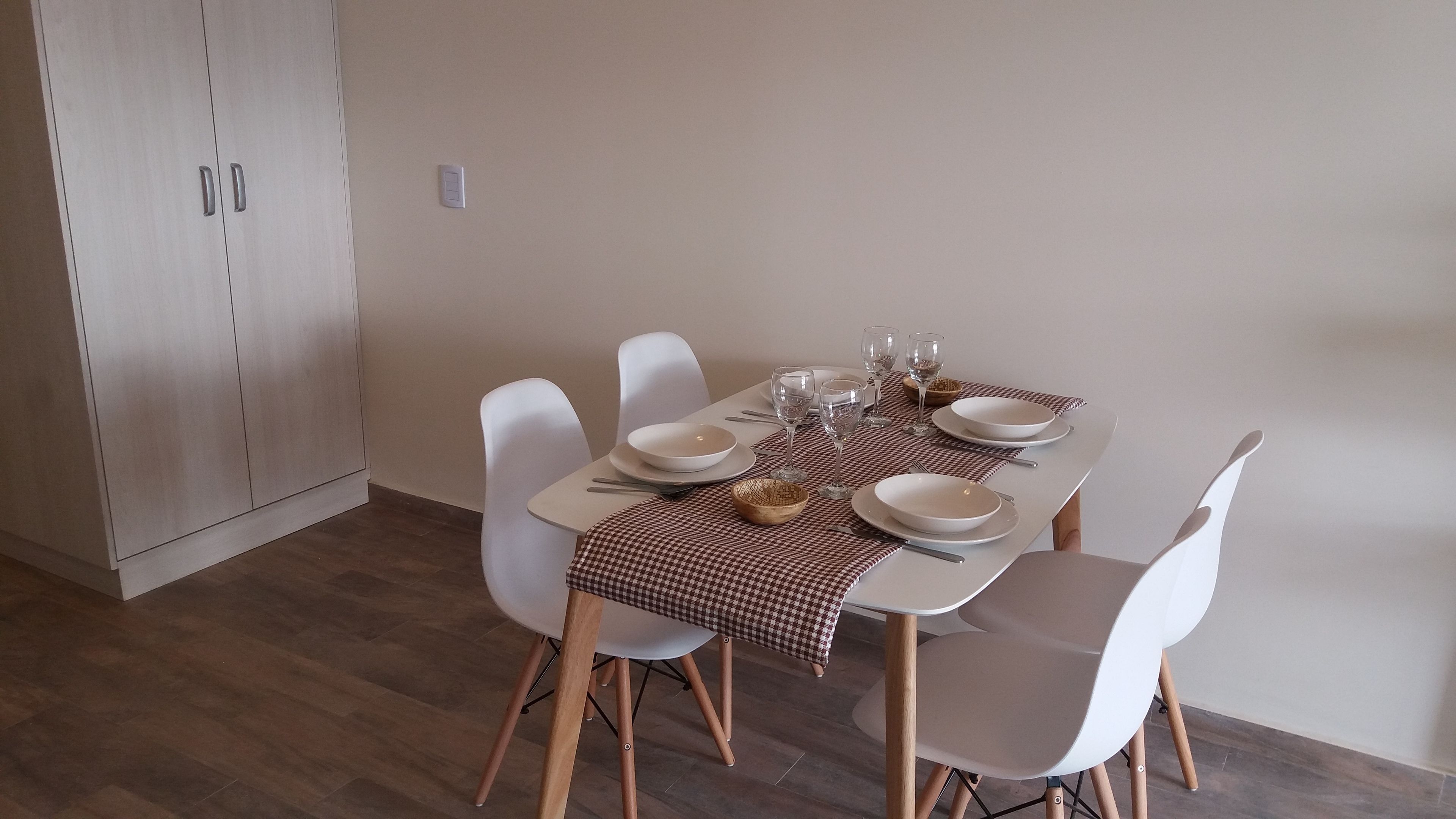Uma área de refeições com uma mesa posta para quatro pessoas, cadeiras brancas e toalha quadriculada.