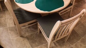 Uma mesa de jantar redonda com jogos americanos verdes, cercada por cadeiras de madeira com assentos acolchoados. Um lustre pendurado está sobre a mesa.