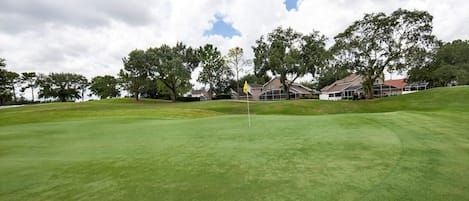 Um campo de golfe com um mastro de bandeira, árvores e casas ao fundo.