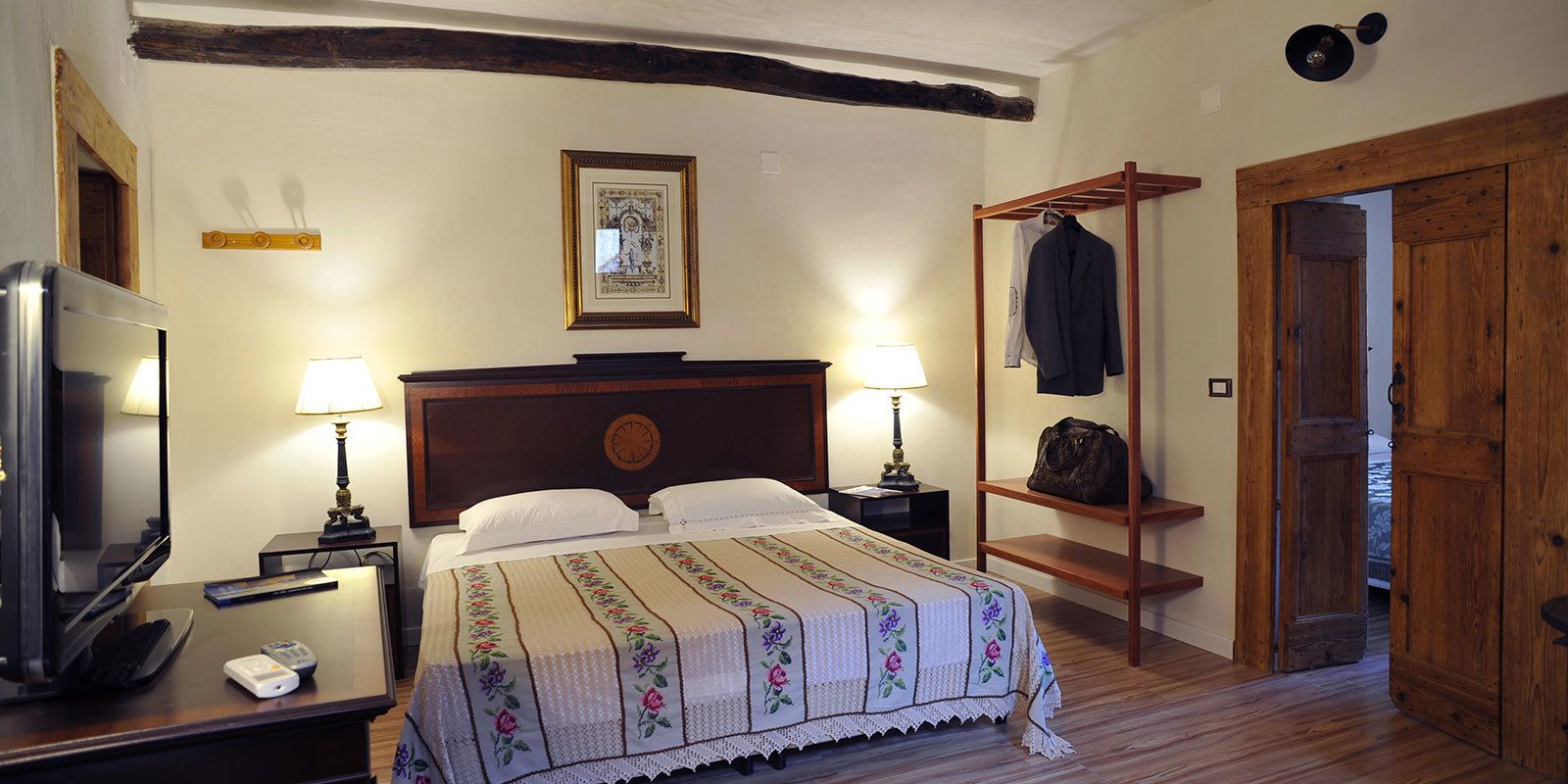 Quarto de hotel com cama, duas luminárias de cabeceira, cabeceira de madeira, um quadro na parede, um cabideiro com um casaco e uma porta de madeira.