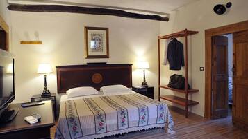 Quarto de hotel com cama, duas luminárias de cabeceira, cabeceira de madeira, um quadro na parede, um cabideiro com um casaco e uma porta de madeira.