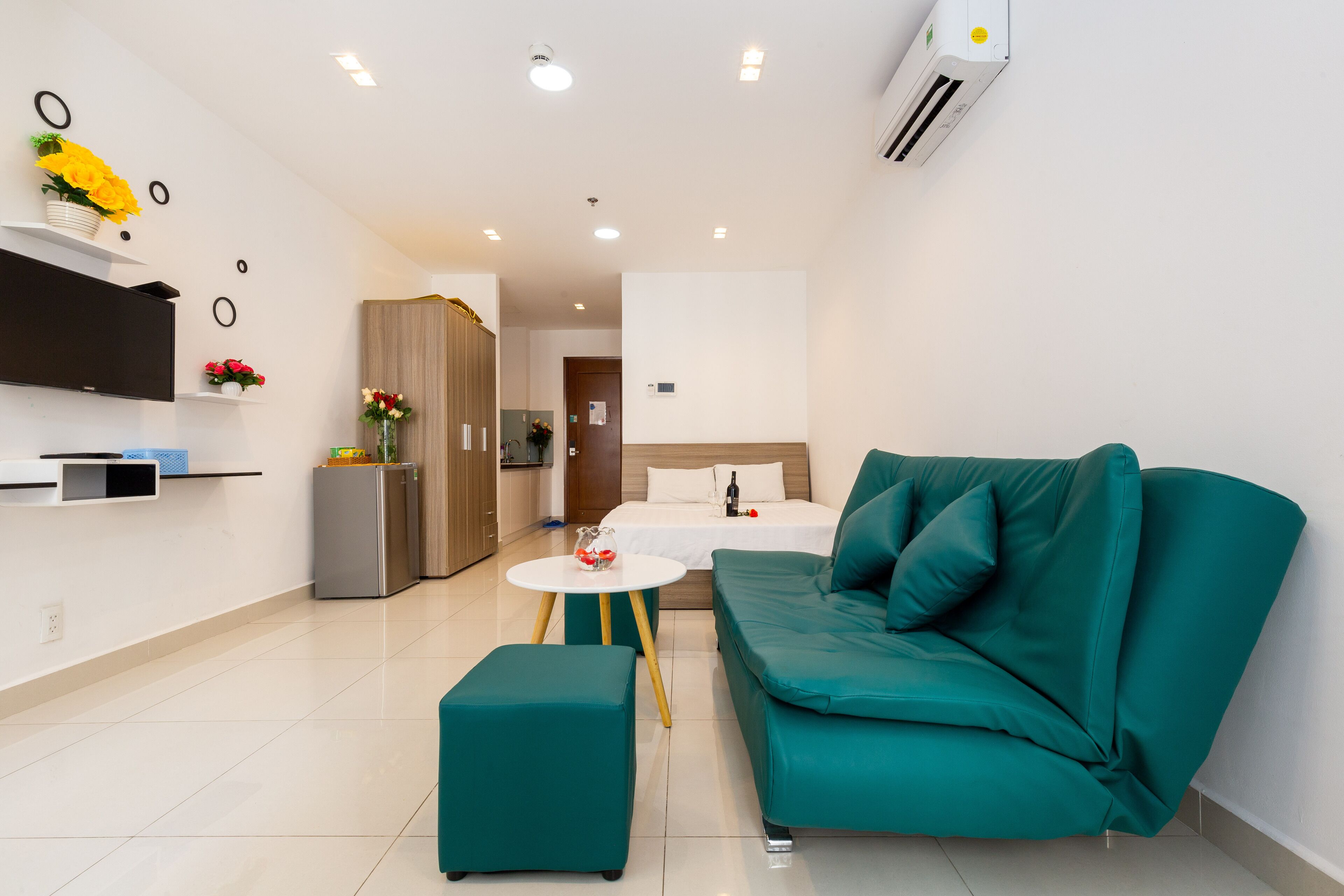 Uma sala de estar moderna com um sofá azul-turquesa, um banquinho coordenado, uma pequena mesa redonda e uma área de cozinha ao fundo.