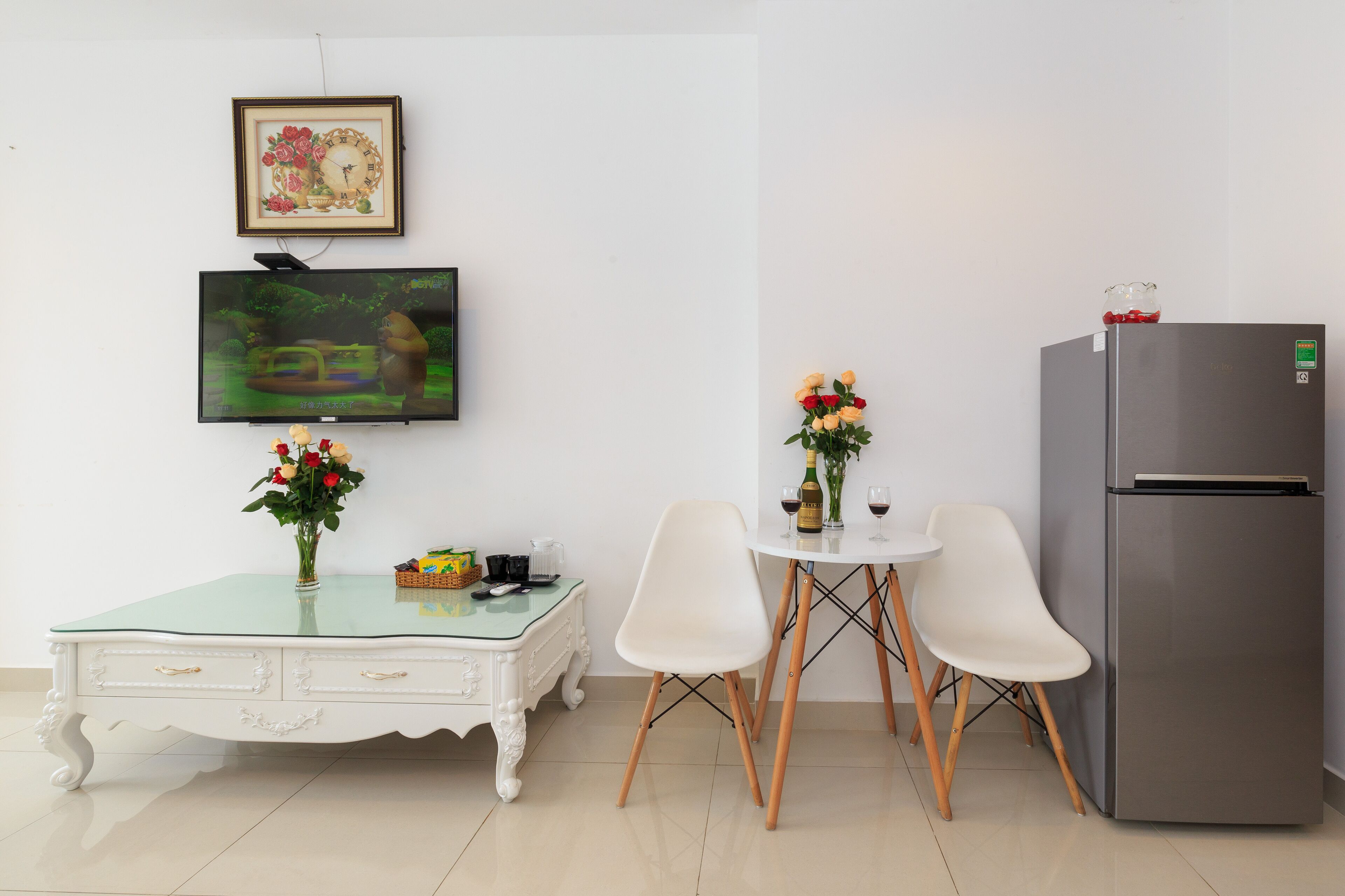 Cozinha moderna com mesa branca, duas cadeiras brancas, mesa de vidro, geladeira, televisão e um quadro na parede.