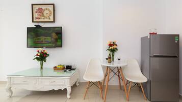 Cozinha moderna com mesa branca, duas cadeiras brancas, mesa de vidro, geladeira, televisão e um quadro na parede.