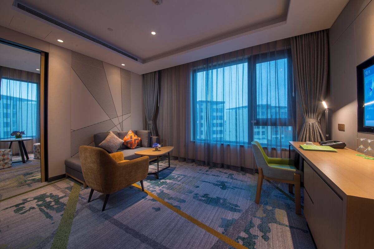 Quarto de hotel com sofá, poltrona, escrivaninha e grandes janelas.
