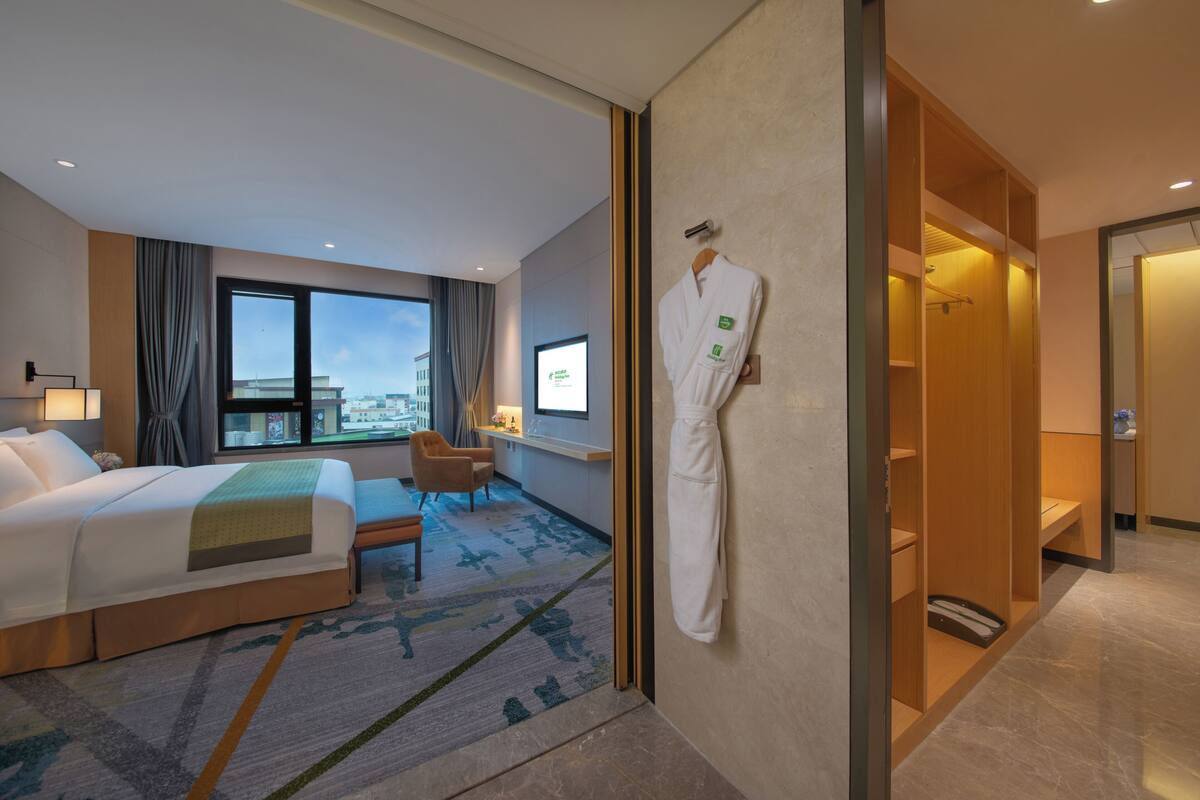 Quarto de hotel com uma cama grande, uma mesa de trabalho, uma cadeira, um roupão pendurado na parede e vista para a cidade pela janela.